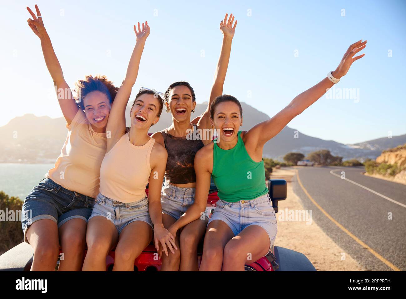 Portrait Of Laughing Female Friends Sitting On Hood Of Open Top Car On ...