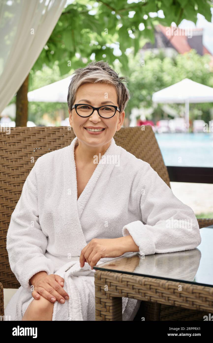 happy middle aged woman in glasses and white robe sitting at rattan table during wellness ...