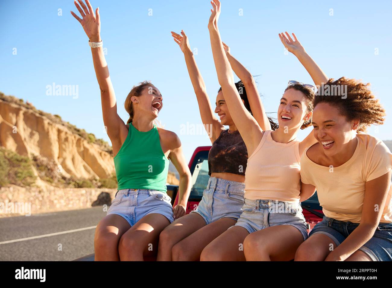 Laughing Female Friends Sitting On Hood Of Open Top Car On Road Trip ...