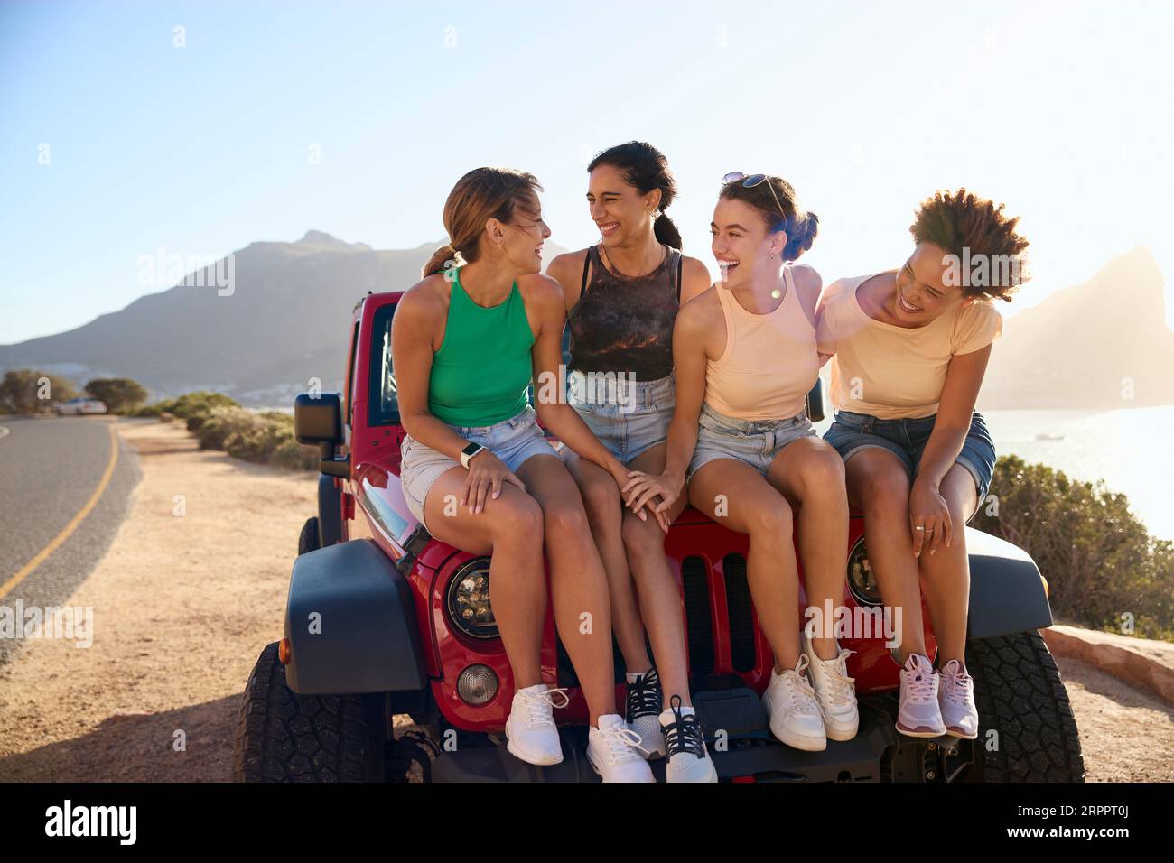 Laughing Female Friends Sitting On Hood Of Open Top Car On Road Trip ...