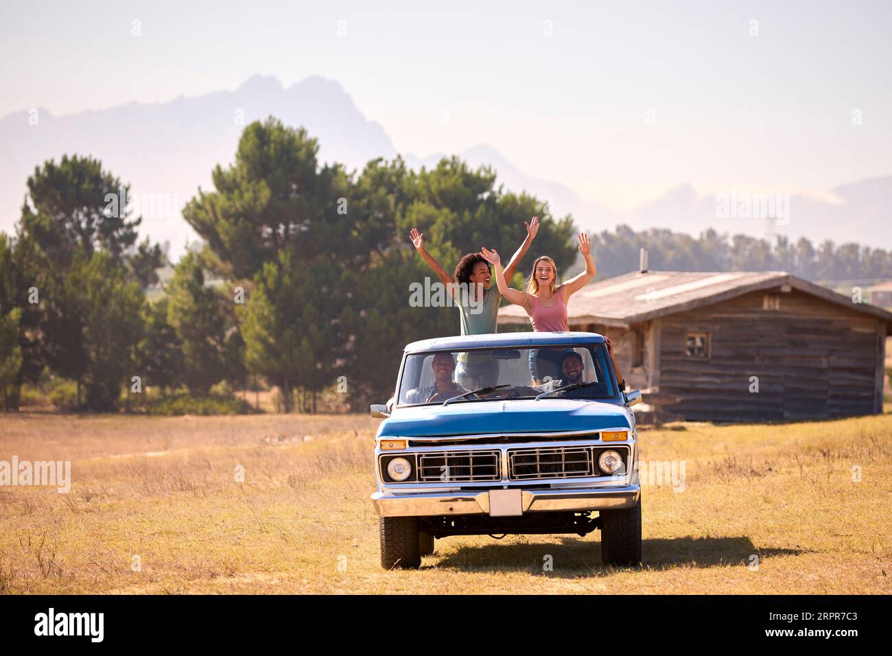 Two Women Standing In Back Of Pick Up Truck As Friends Enjoy Road Trip ...