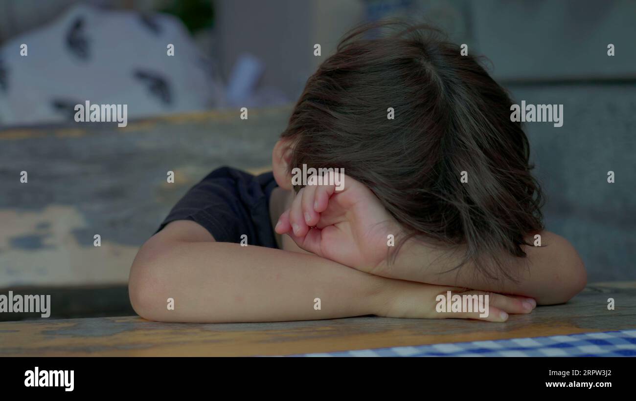 Bored child covering face with arms leaning on table feeling boredom ...