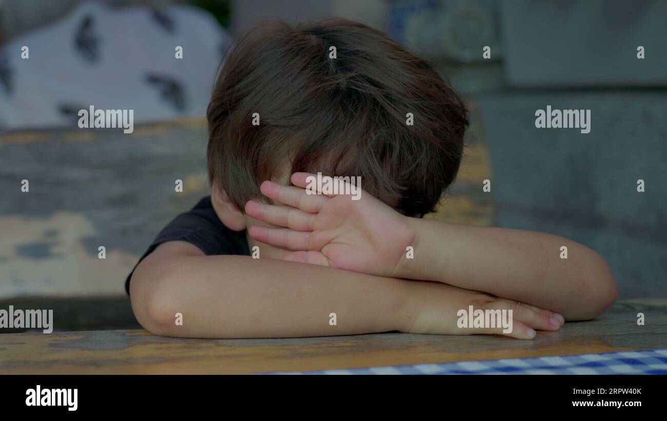 Bored child covering face with arms leaning on table feeling boredom ...
