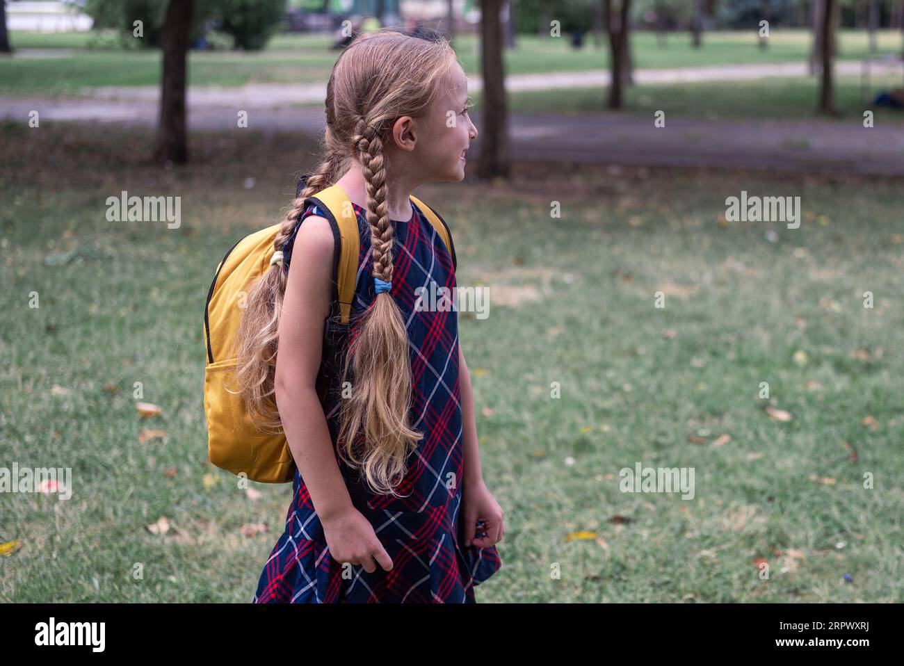 Cute smiling schoolgirl with backpack looks at path in green park ...
