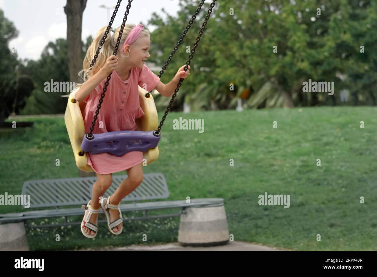 A joyful girl in a pink dress swings with delight in an urban park's ...