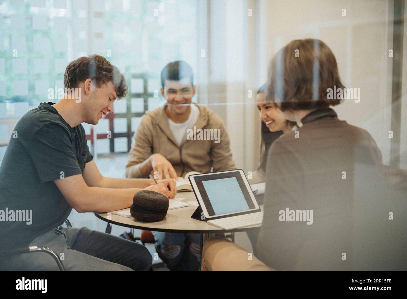 Happy young students studying at table together seen through glass in ...