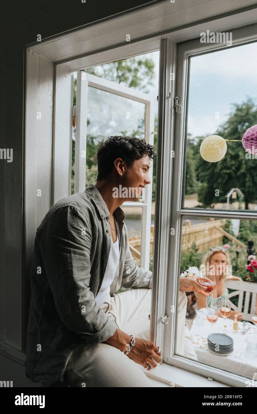 Smiling young man sitting on window sill while looking at friends ...