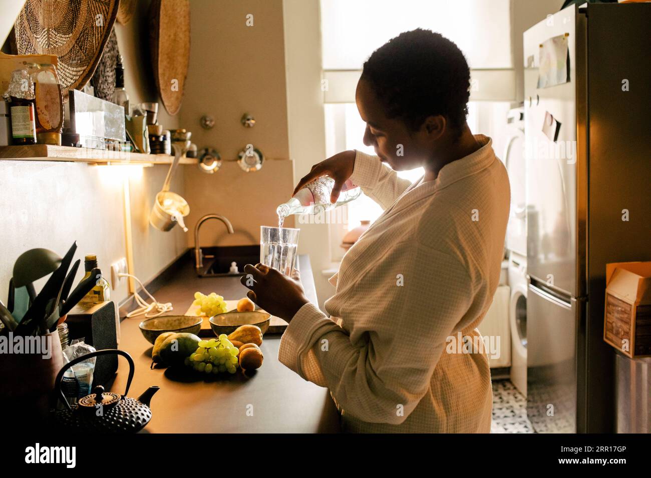 Side view of woman wearing bathrobe pouring water in glass at kitchen ...