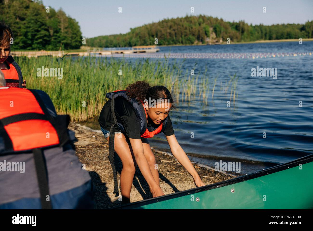 Girl wearing life jacket while holding kayak near lake at summer camp ...