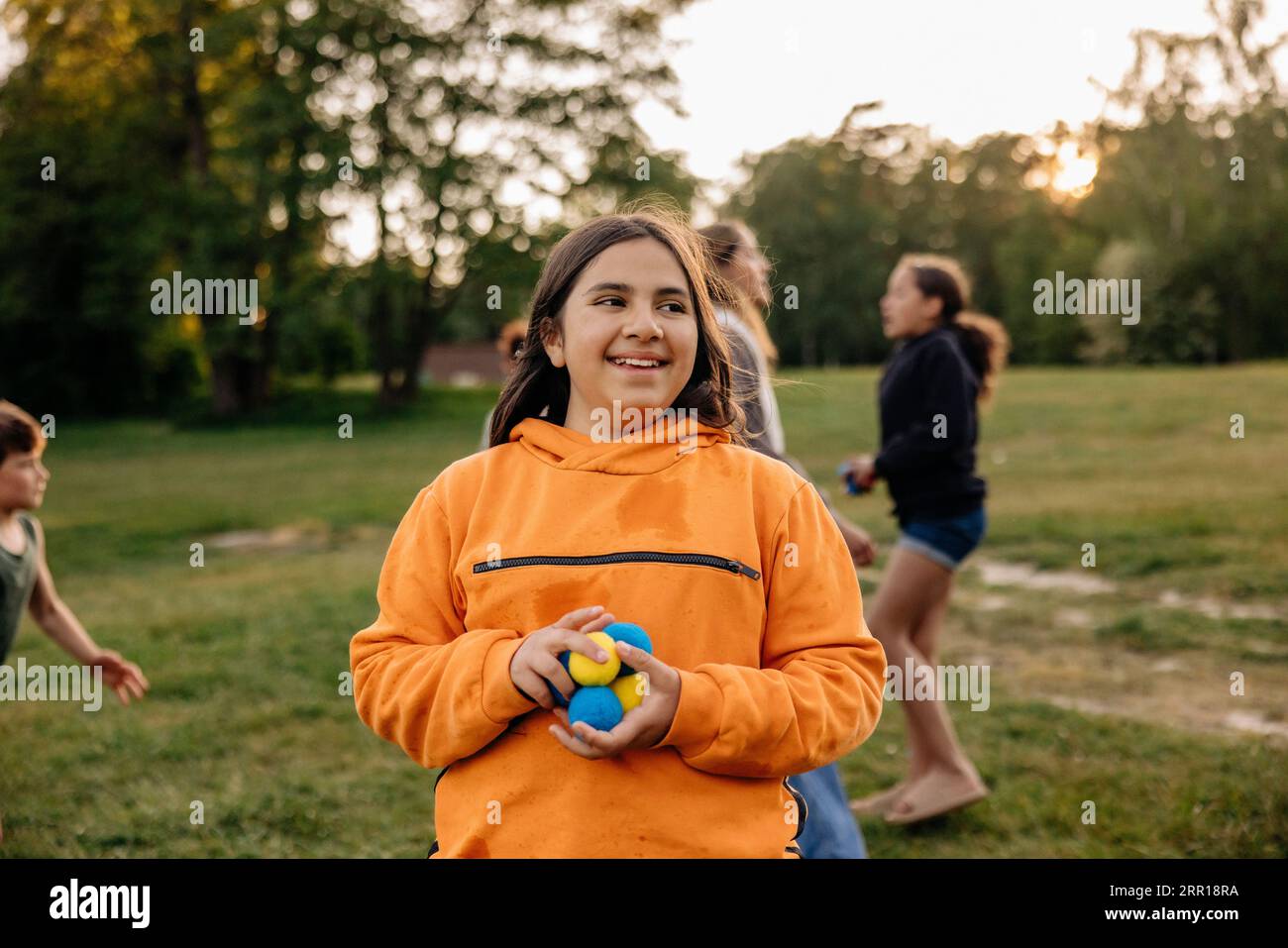 Smiling girl holding blue and yellow balls while standing in playground ...