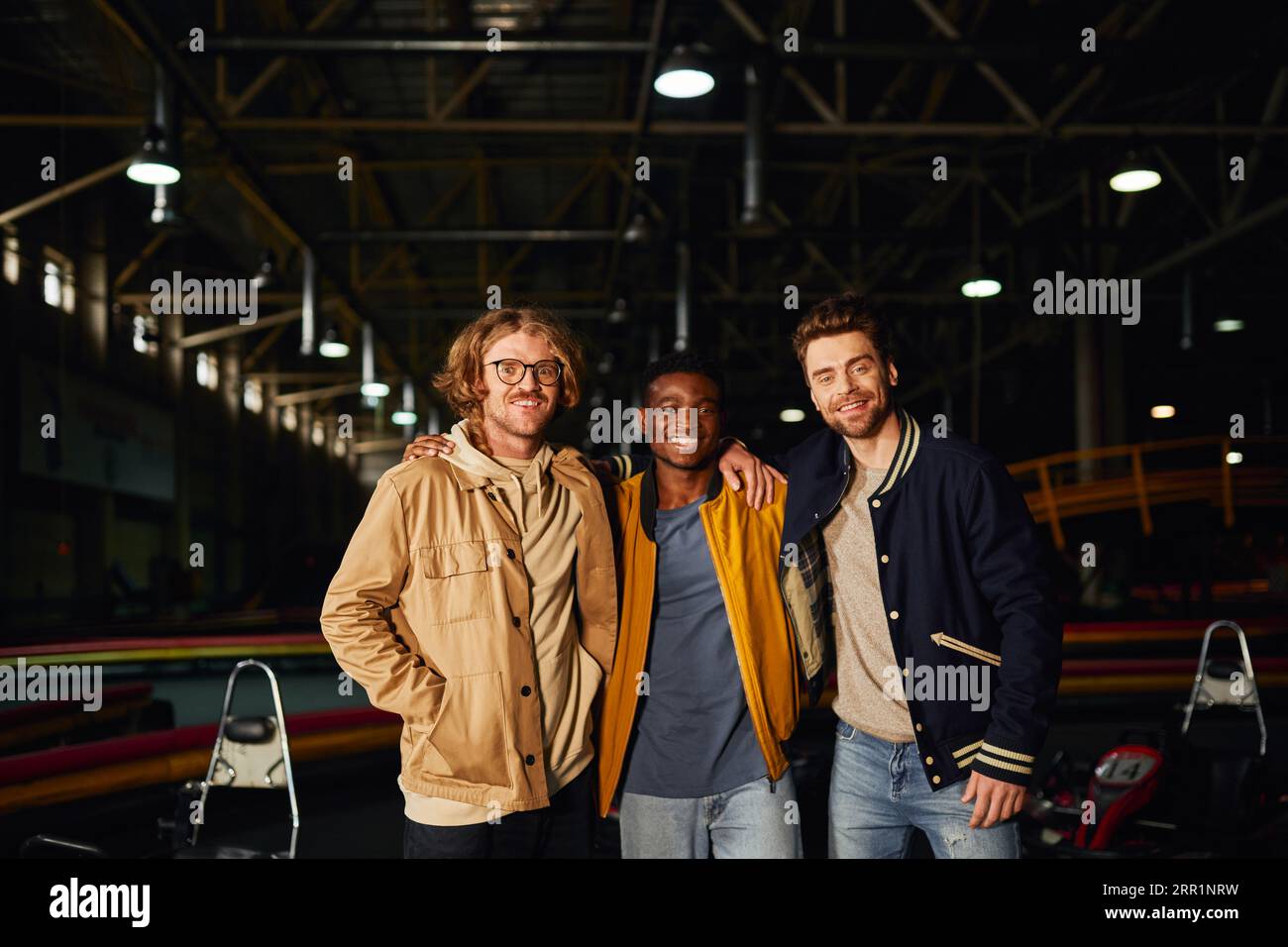 multicultural male friends smiling and standing near racing cars inside ...