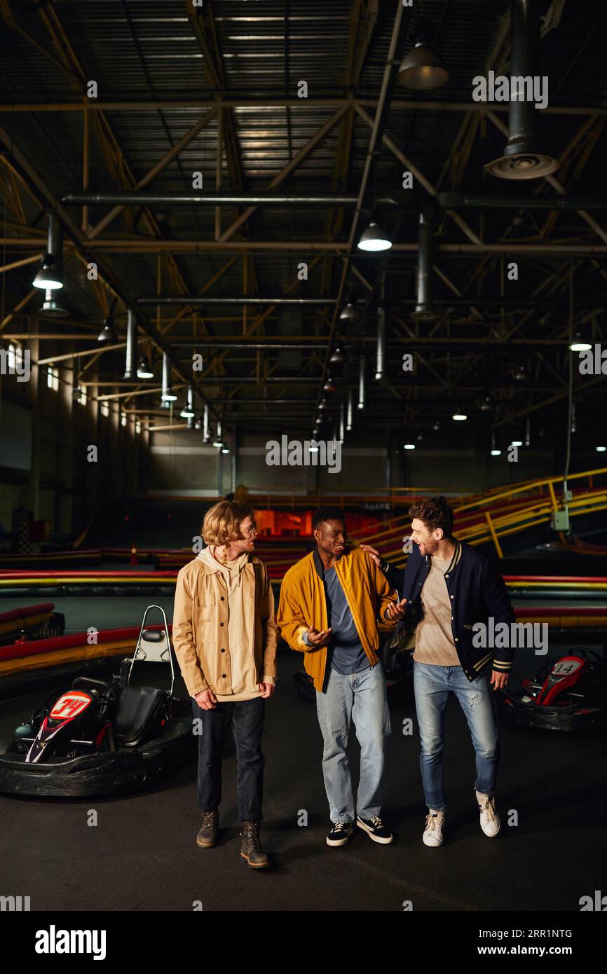 cheerful diverse friends chatting and walking near racing cars inside ...