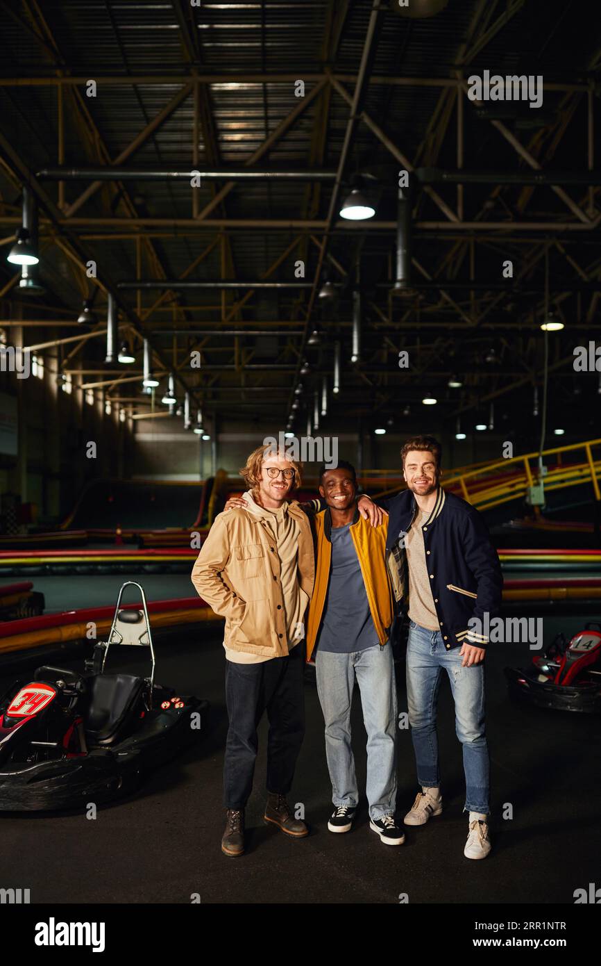 multicultural male friends smiling and standing near racing cars inside ...