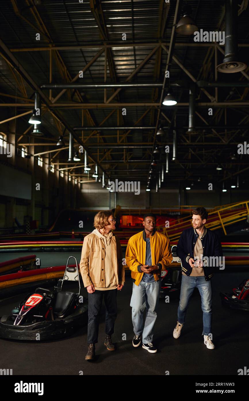 cheerful diverse friends chatting and walking near racing cars inside ...