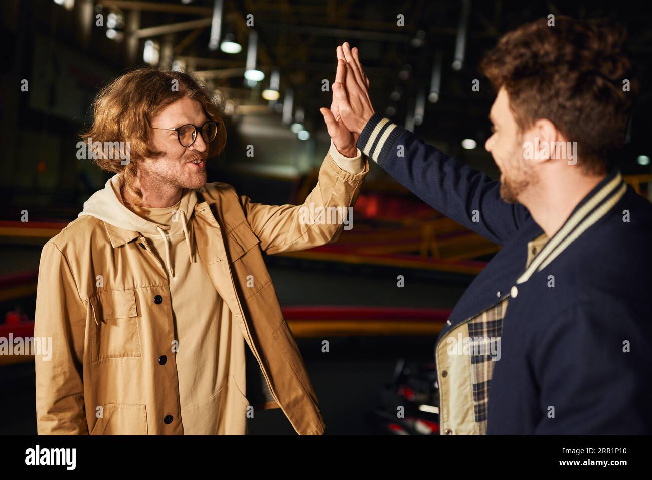 cheerful man in glasses giving high five to friend inside of karting ...