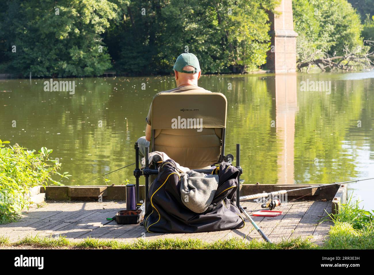 Rear view of man sitting isolated by UK pool/lake fishing in the warm ...