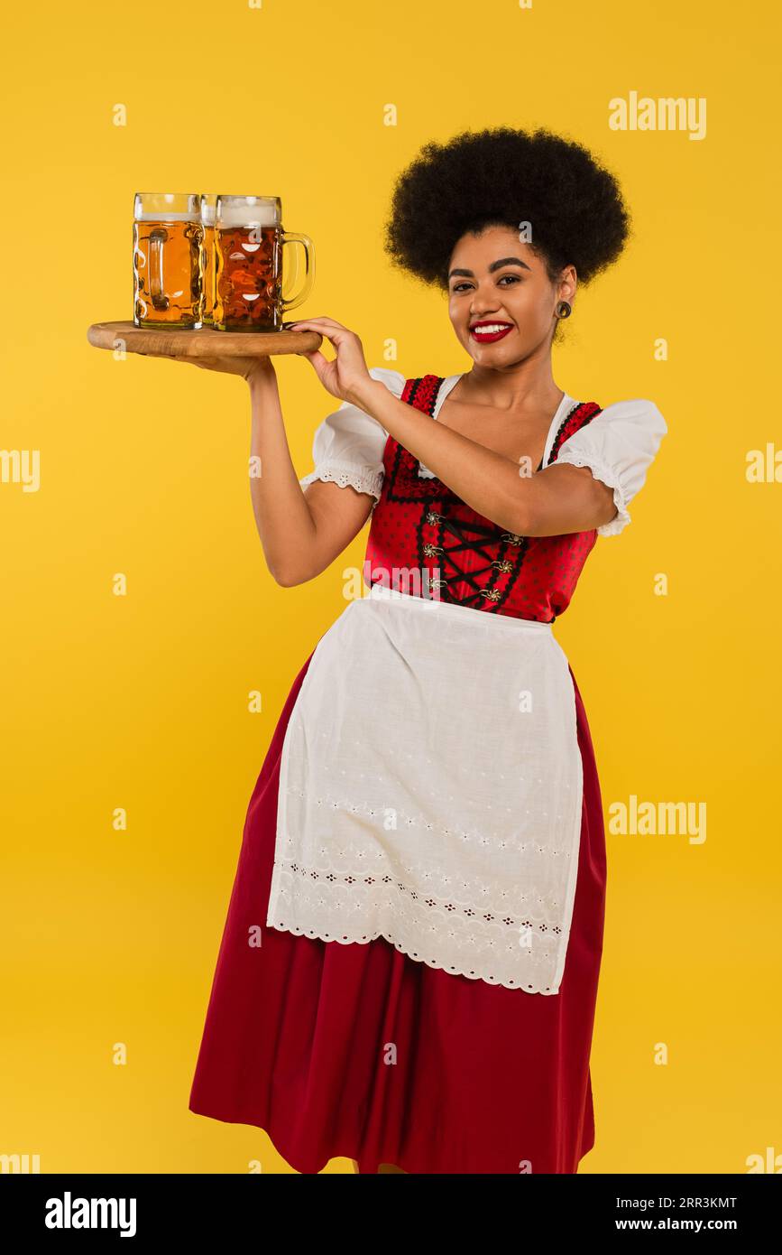 joyful african american bavarian waitress in dirndl serving beer mugs ...
