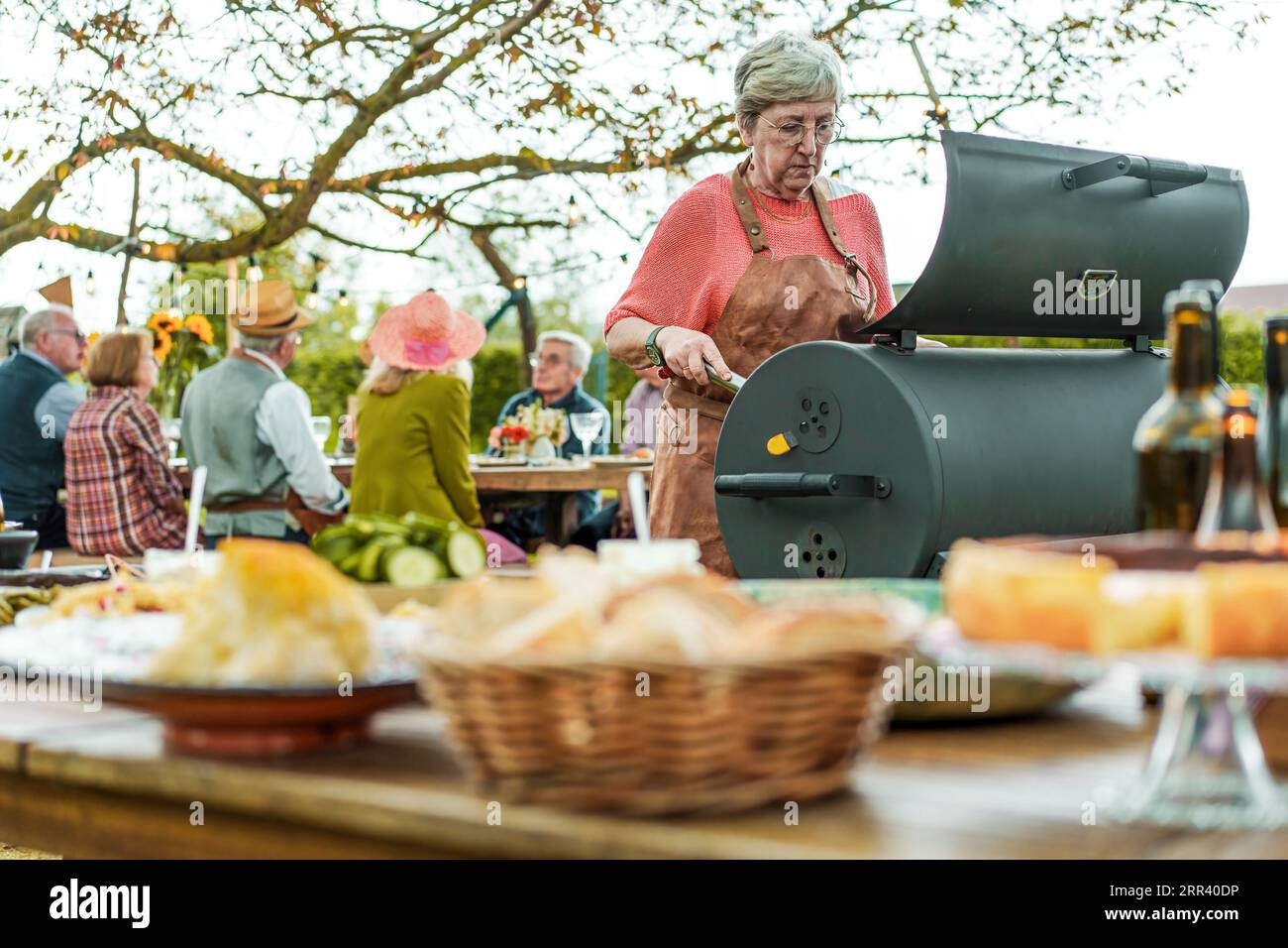 Senior woman with gray hair preps BBQ in the countryside. Blurred ...