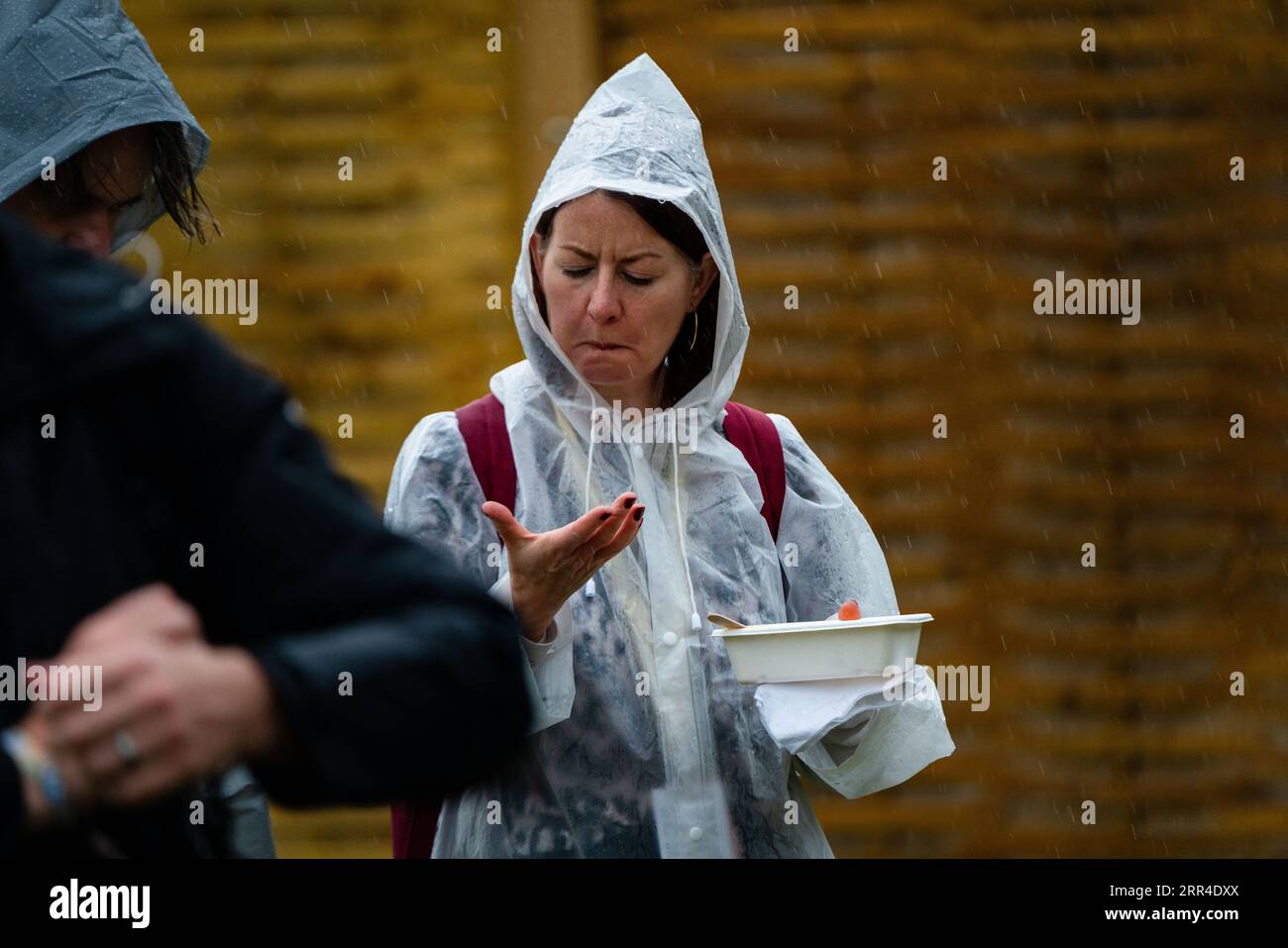 A woman in a plastic mac eating fast food from a tray in the rainstorm ...