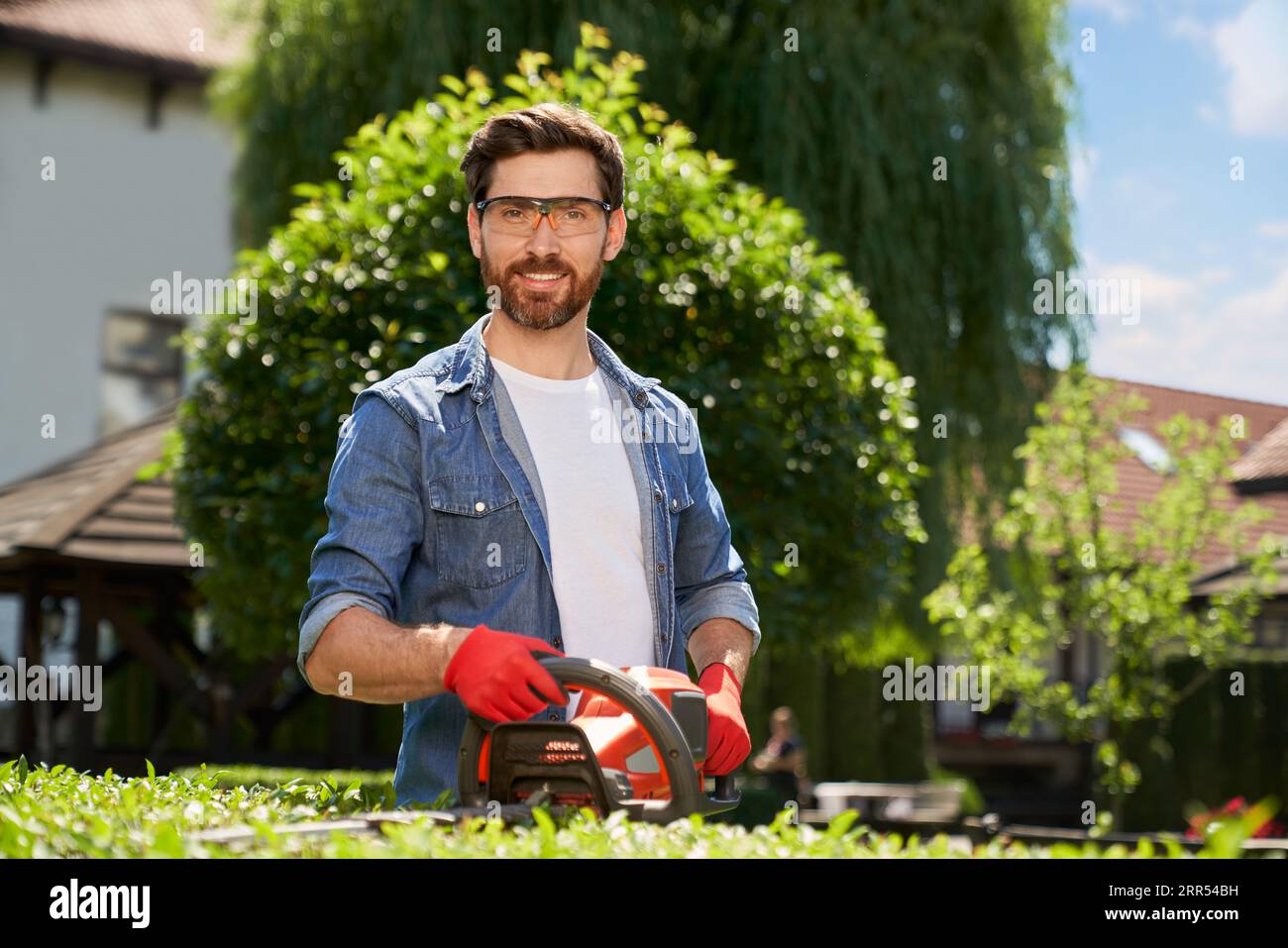 Smiling male landscaper in safety glasses cutting bush with hedge ...