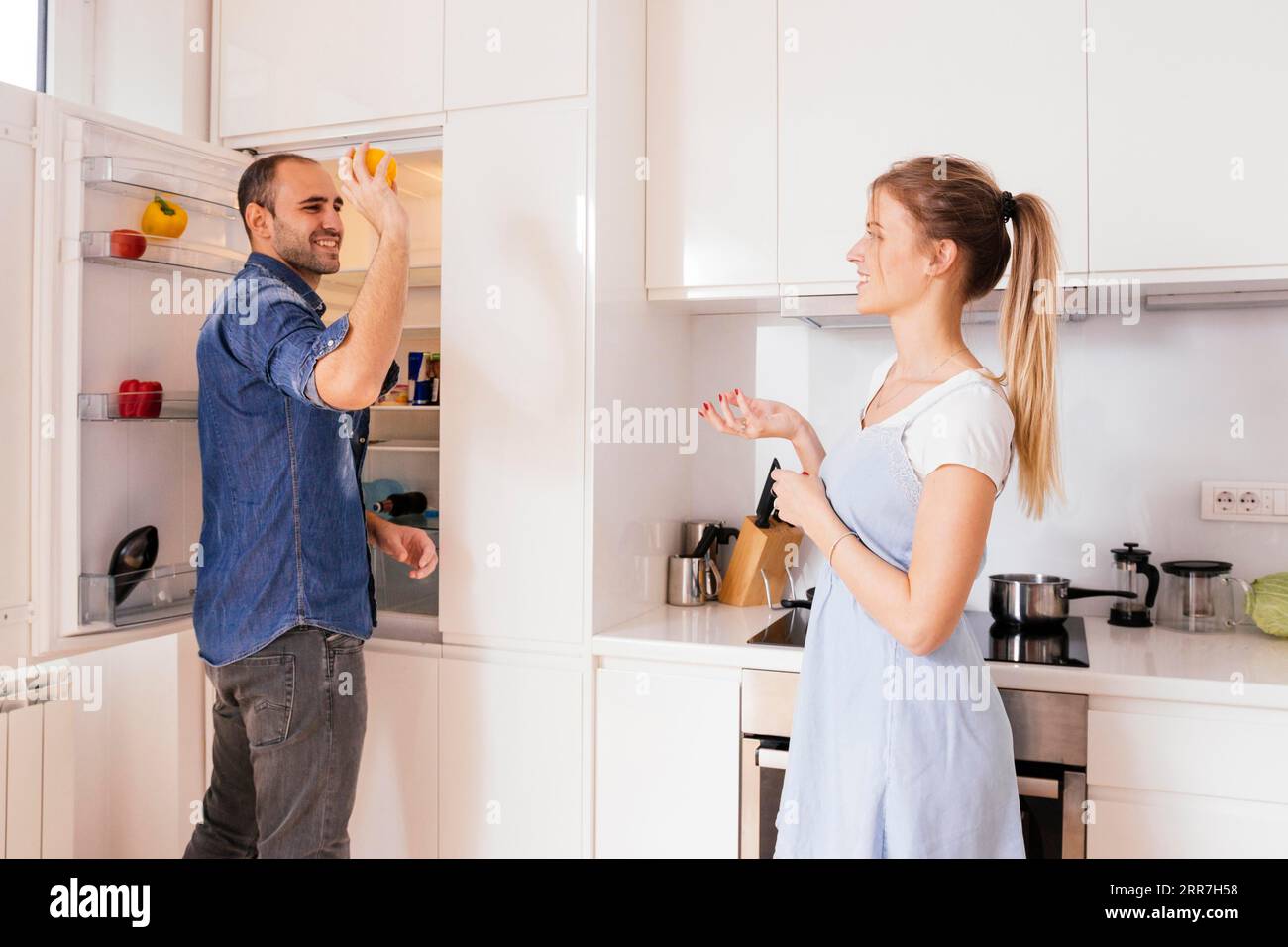 Smiling young man standing near open refrigerator throwing vegetable ...