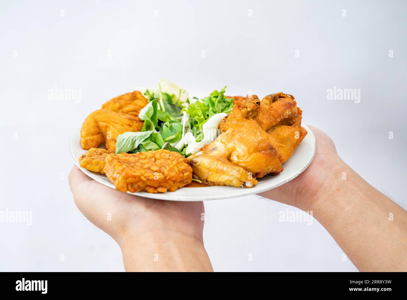 A human hand holding a plate with pecel ayam, fried tempe, and tofu ...