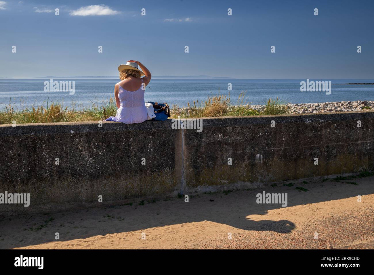 Lone figure of woman sitting on a seewall adjusting her hat in the ...