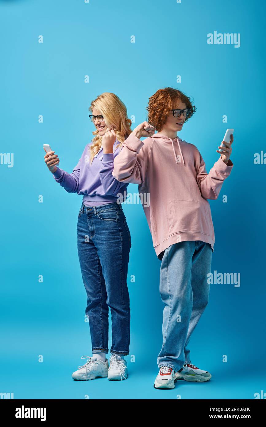 happy and trendy teenage friends in eyeglasses and using smartphones and showing win gesture on ...