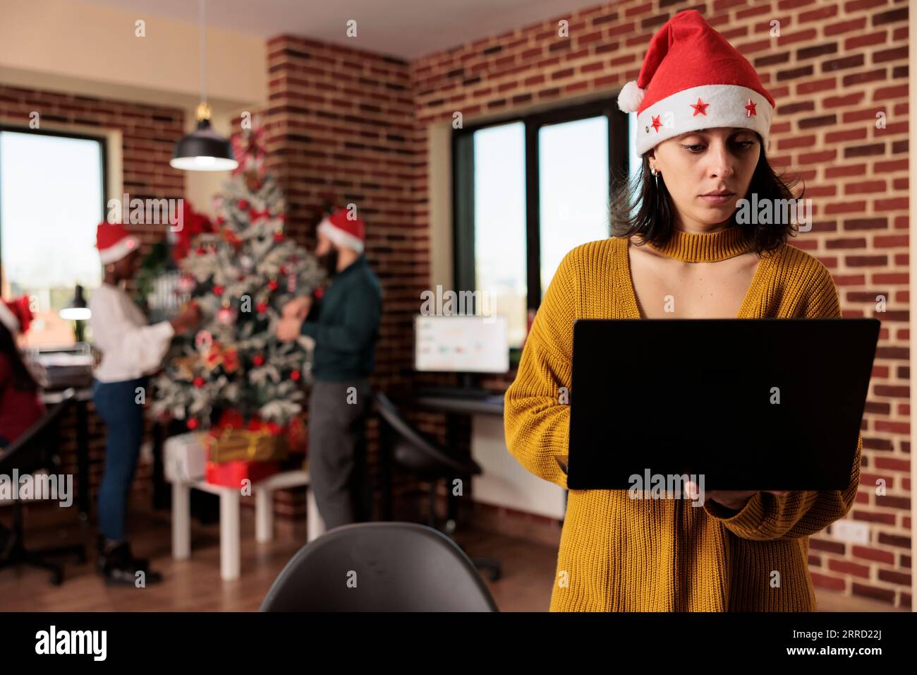 Caucasian woman employee wearing santa claus hat standing with laptop ...