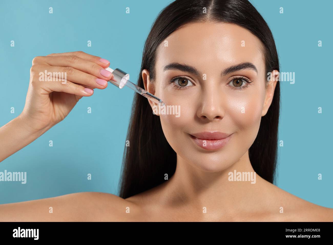 Beautiful young woman applying serum onto her face on light blue ...