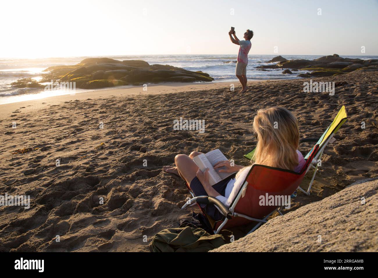 Mature woman reads book in beach chair as man takes photo with tablet ...
