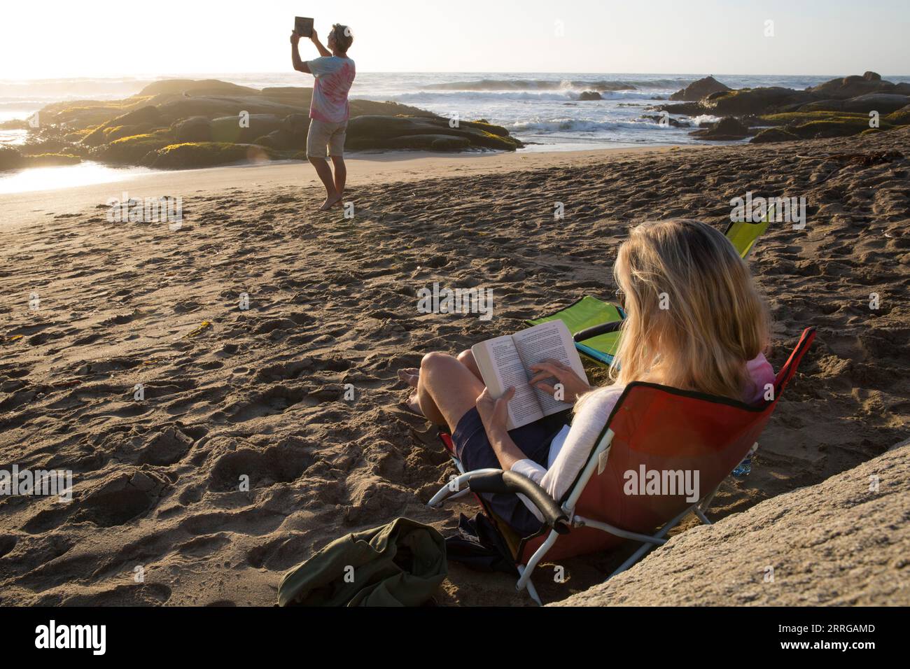 Mature woman reads book in beach chair as man takes photo with tablet ...