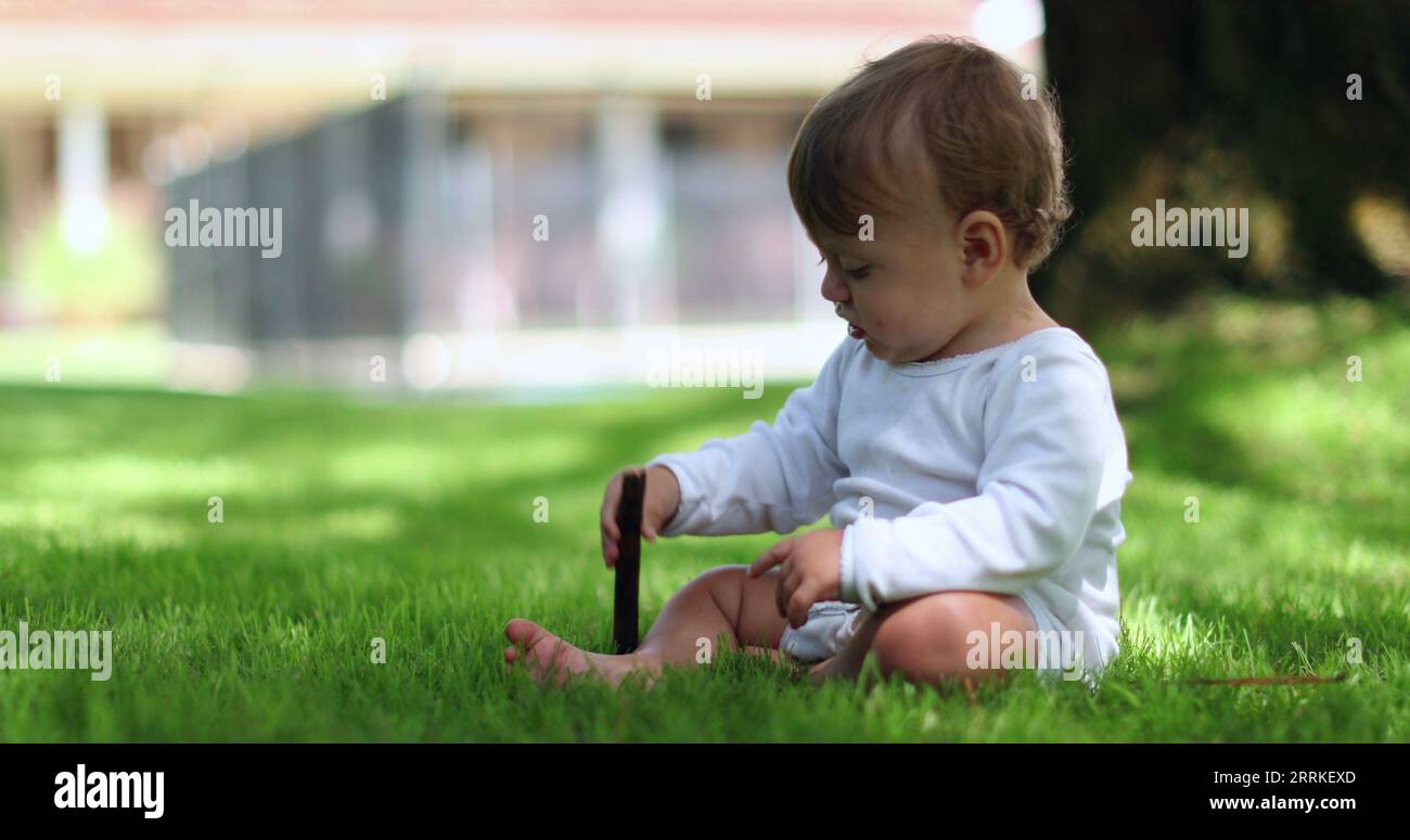 Adorable baby sitting outside in home lawn holding stick. Cute infant ...
