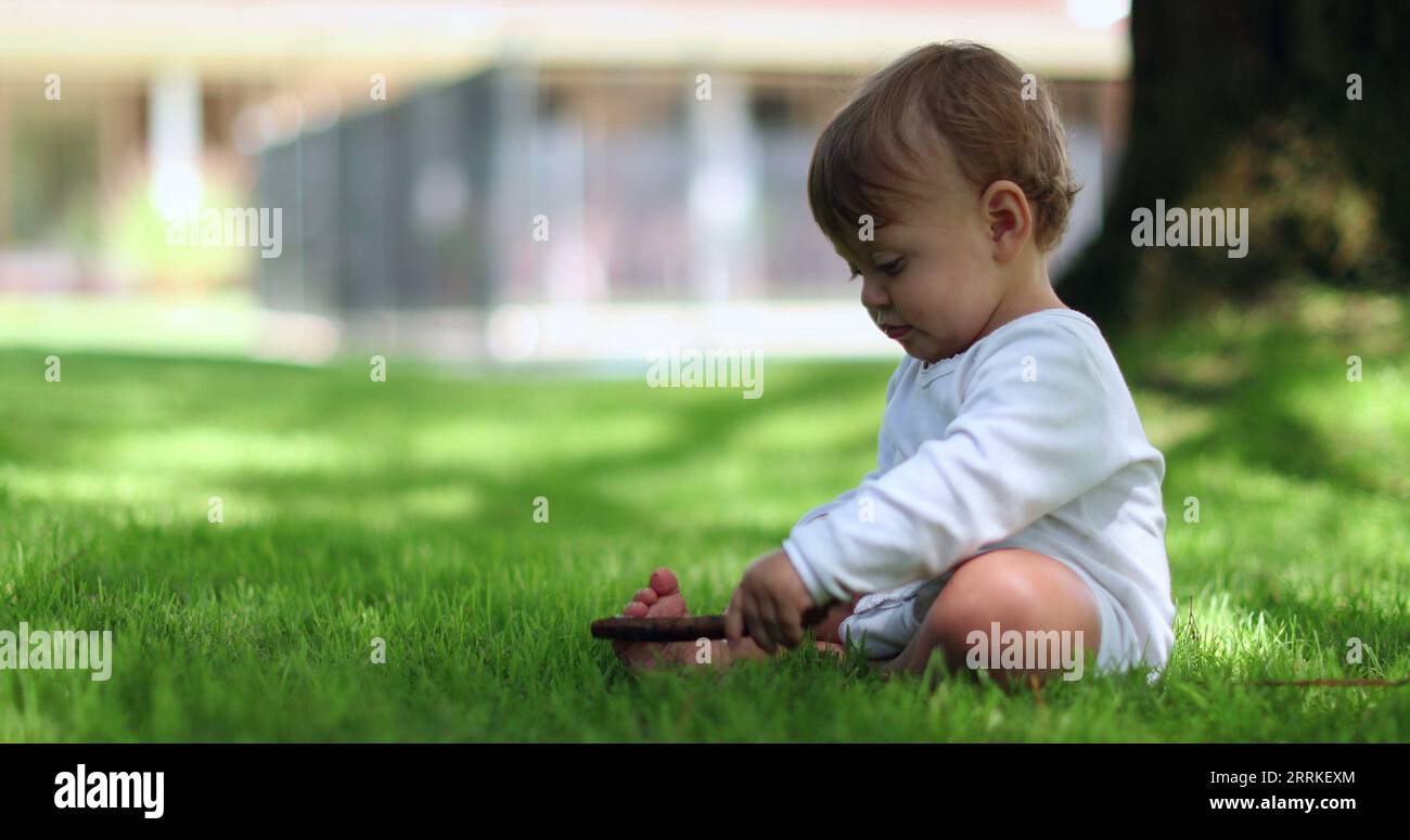 Adorable baby sitting outside in home lawn holding stick. Cute infant ...