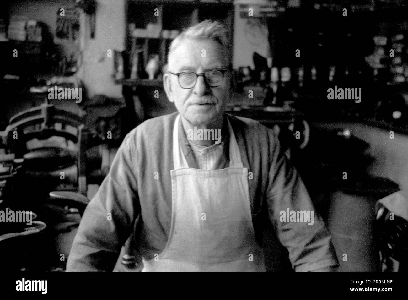 England. c.1960 – A cobbler posing for a photograph in his workshop. He ...