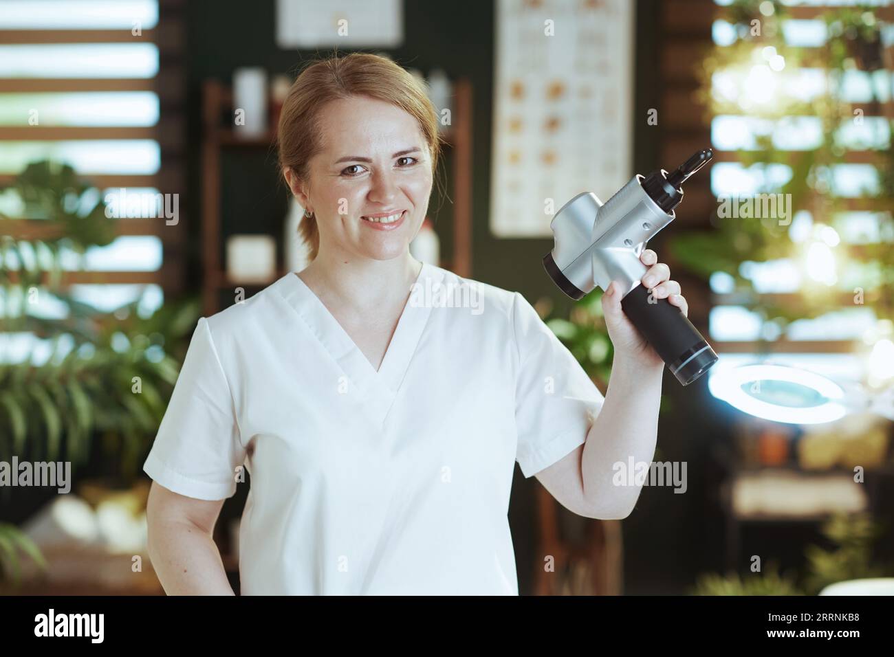 Healthcare time. smiling female medical massage therapist in massage cabinet with massage pistol ...