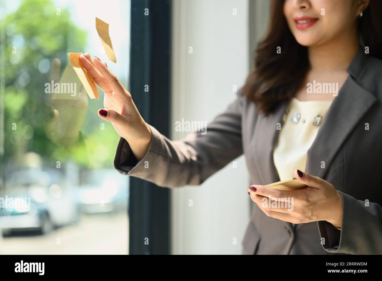 Smiling businesswoman sticking adhesive notes on glass window, planning ...