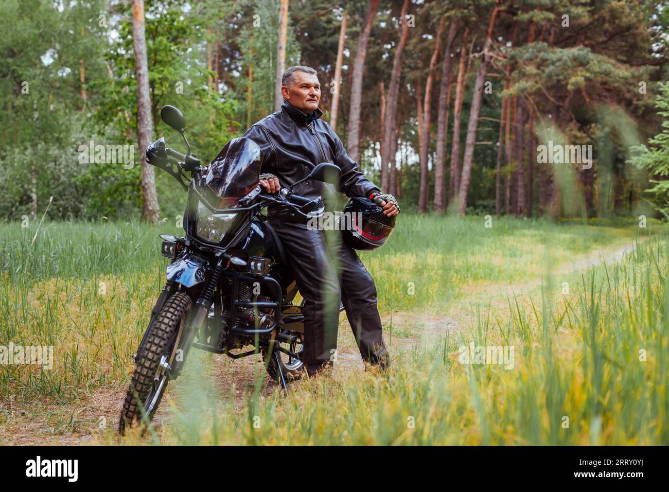 Elderly biker leaning on motobike in summer forest. Man in leather ...