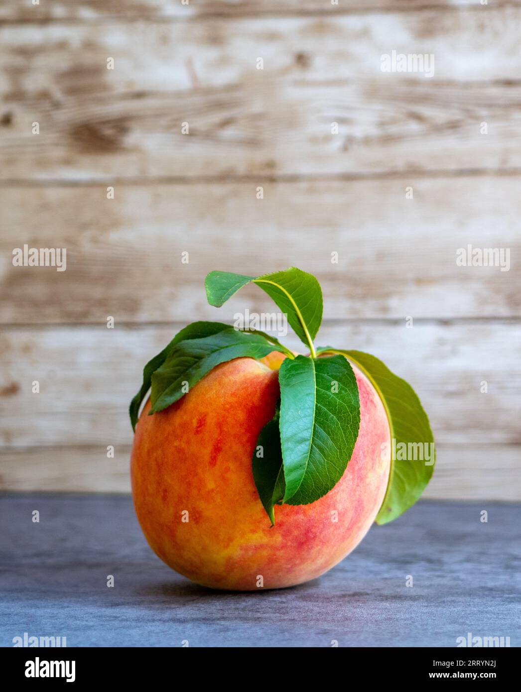A single peach with leaves rests on a slate table with a rustic wood ...
