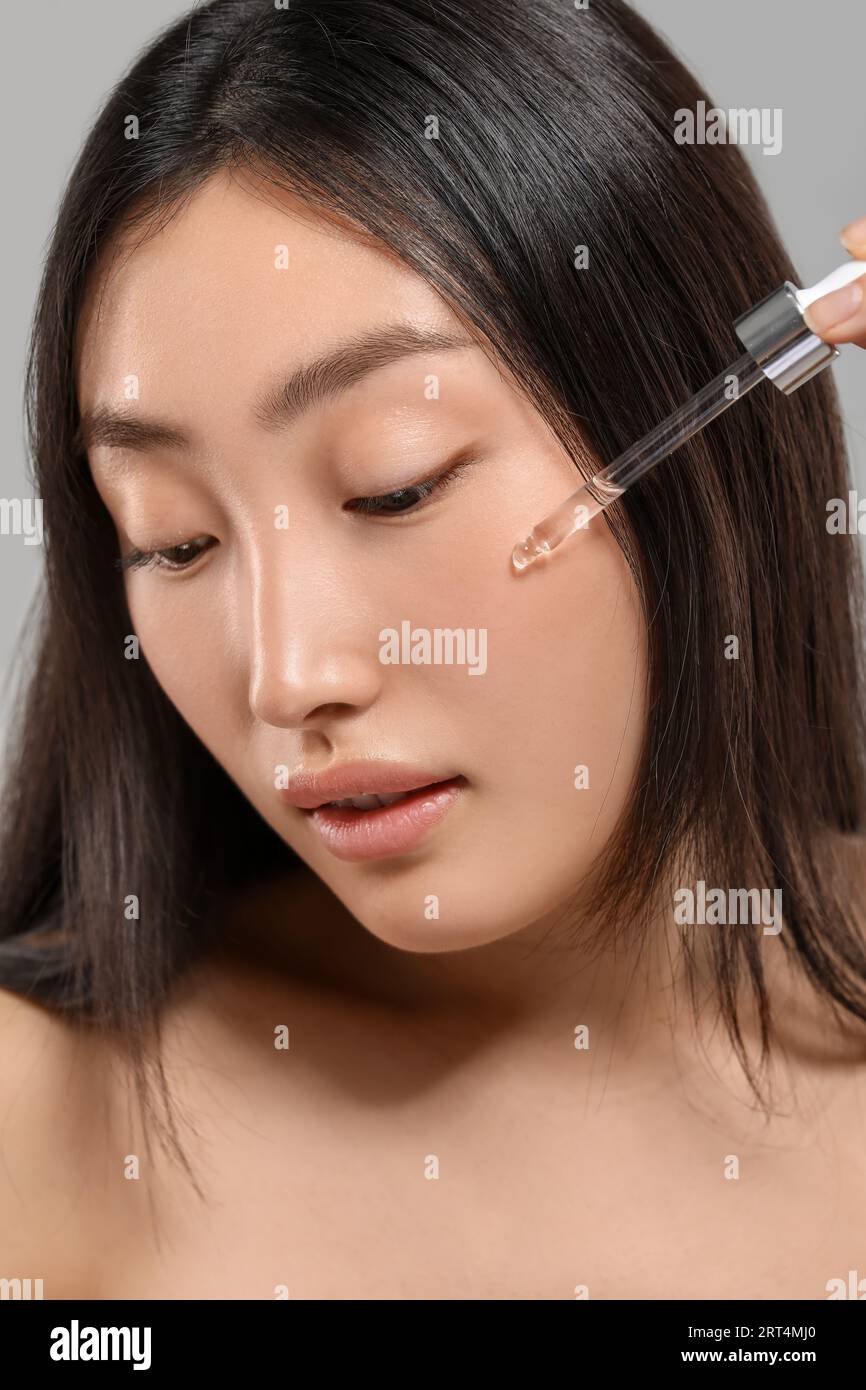 Beautiful young woman applying cosmetic serum onto her face on grey ...