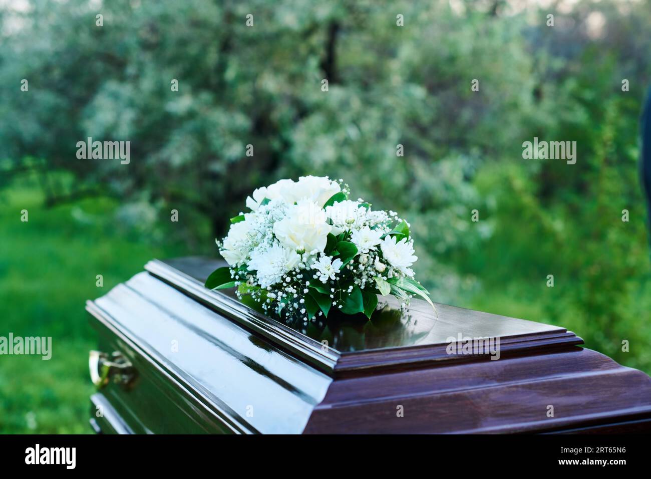 Wooden coffin covered with heavy lid with bunch of white flowers standing against green trees ...