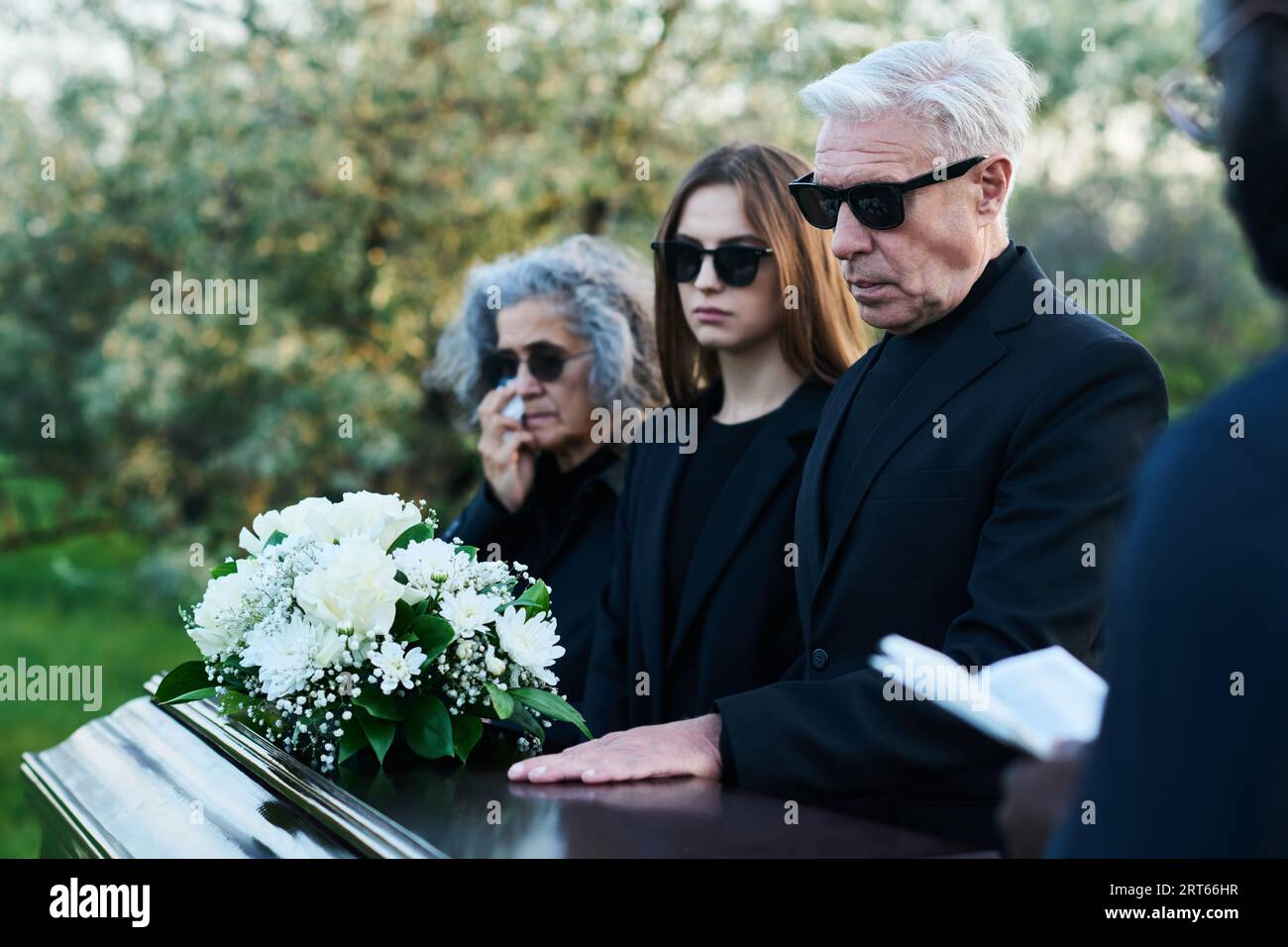 Mature man in sunglasses and mourning attire keeping hand on lid of ...