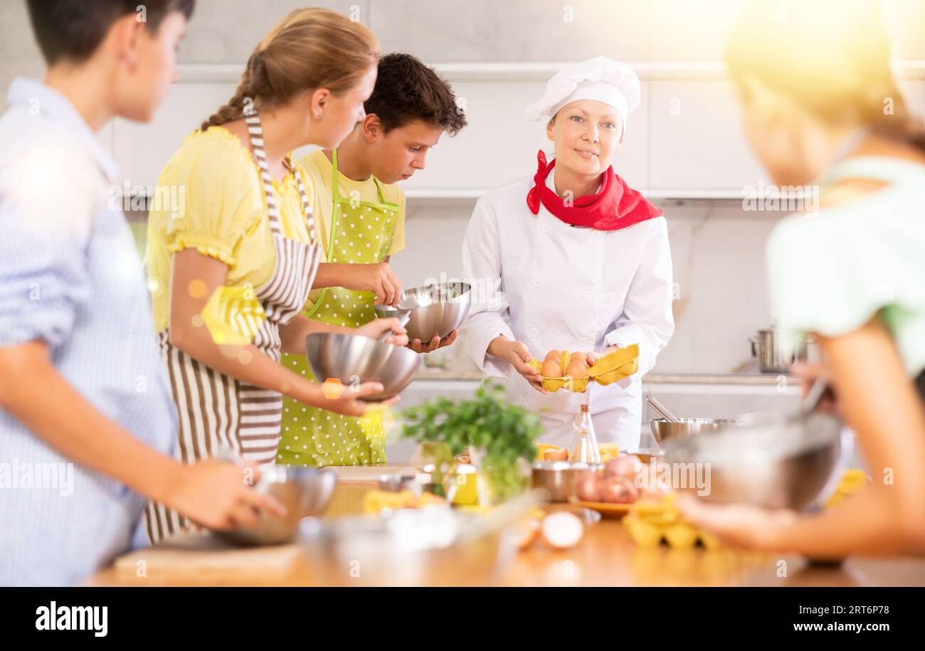 Friendly female chef in white uniform giving culinary lesson to tweens ...