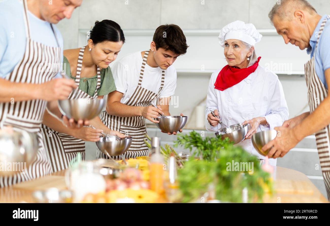 Smiling old woman chef of cooking course teaching attendees how to mix ...