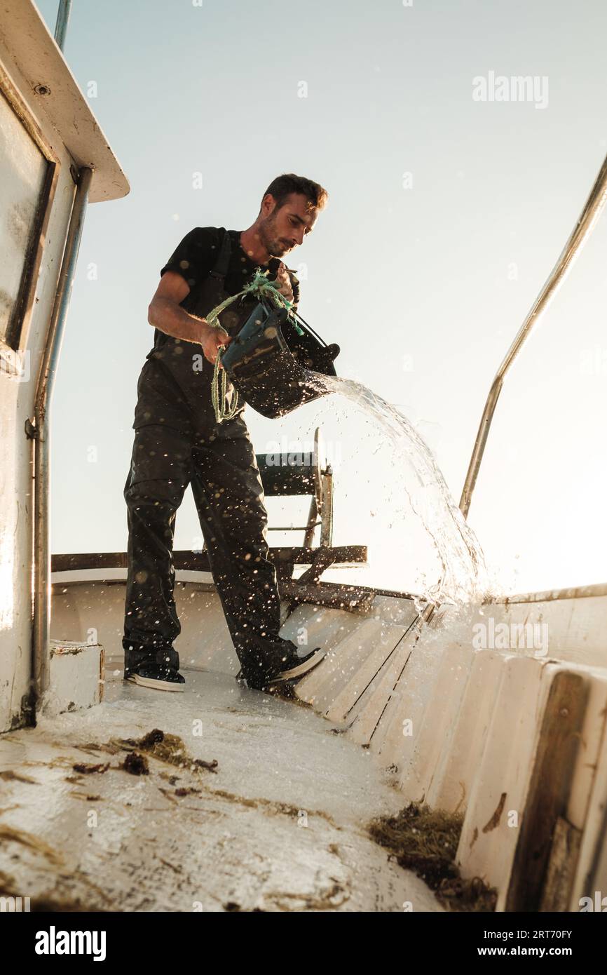 Low angle of bearded male fisher pouring water out of bucket on dirty ...