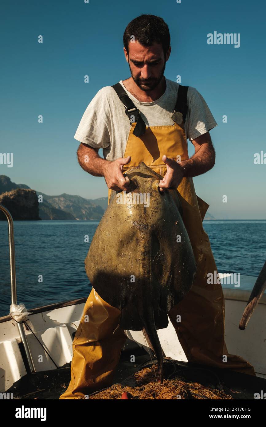 Male fisher with sea stingray floating on boat in sea in sunny summer ...