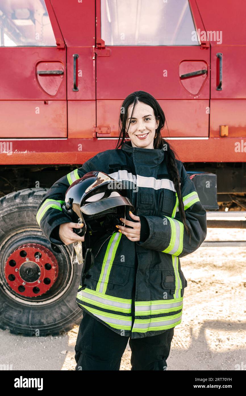 Young positive female firefighter with black pigtail holding helmet while wearing uniform ...