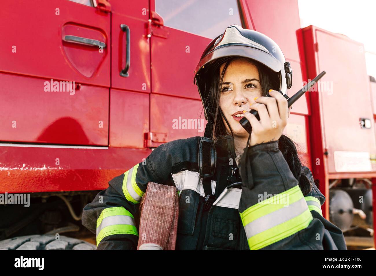 Young female firefighter in helmet carrying hose and communicating on ...