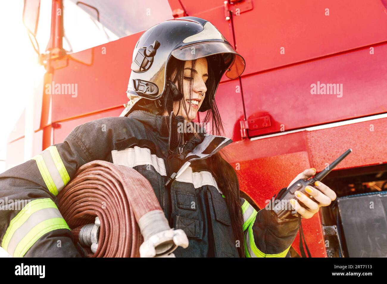 Young female firefighter in helmet carrying hose with walkie talkie ...