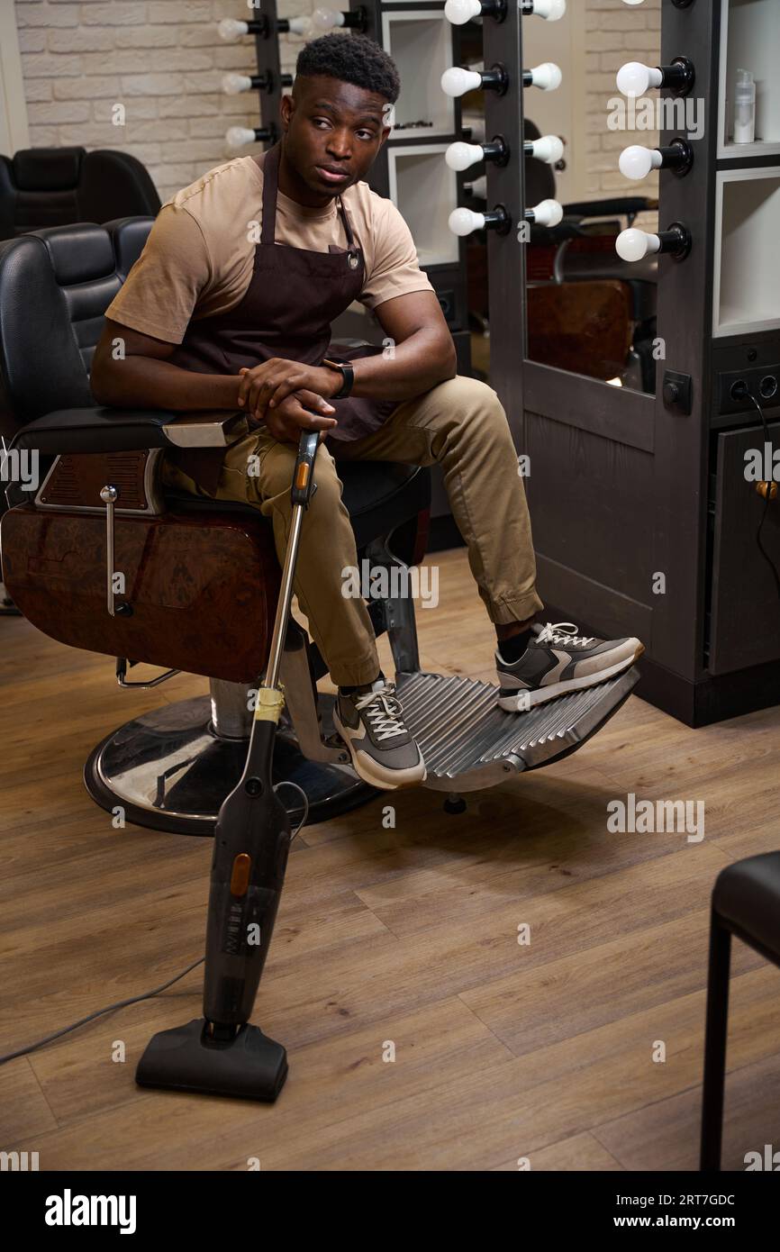 African American barber in work apron resting in hairdressers chair ...